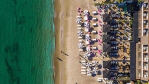 Vista aérea de um hotel à beira-mar com fileiras de espreguiçadeiras e guarda-sóis, uma praia arenosa e um edifício com varandas com vista para o oceano.