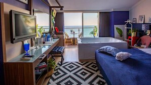 Quarto de hotel moderno com uma janela ampla com vista para o mar, televisão de tela plana, pequena cozinha, banheira e cama com manta azul.