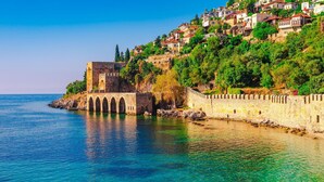Um edifício histórico de pedra com arcos à beira de um mar azul límpido, rodeado por vegetação exuberante e edifícios residenciais numa encosta.