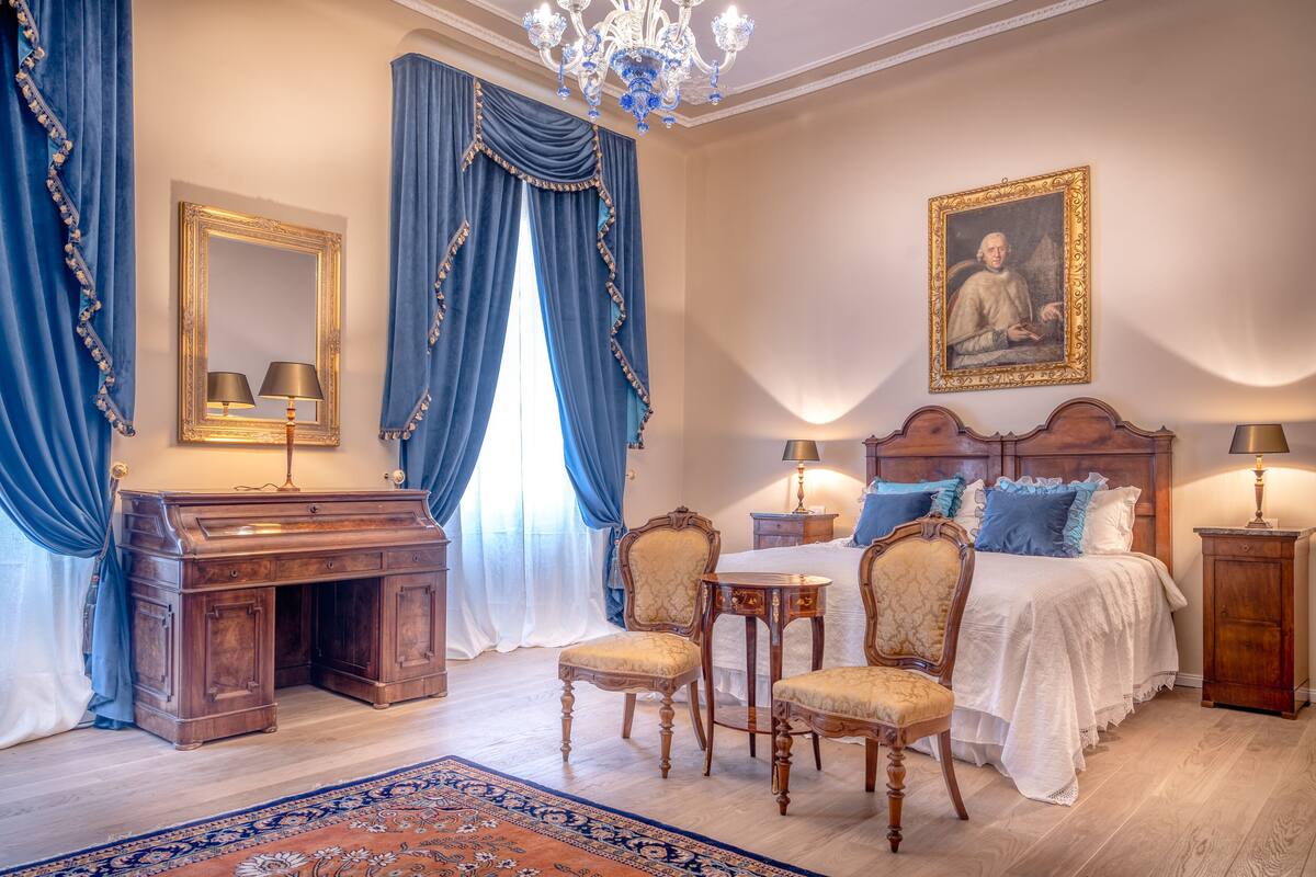 Quarto clássico com escrivaninha de madeira, duas cadeiras, cama com almofadas azuis e um lustre.