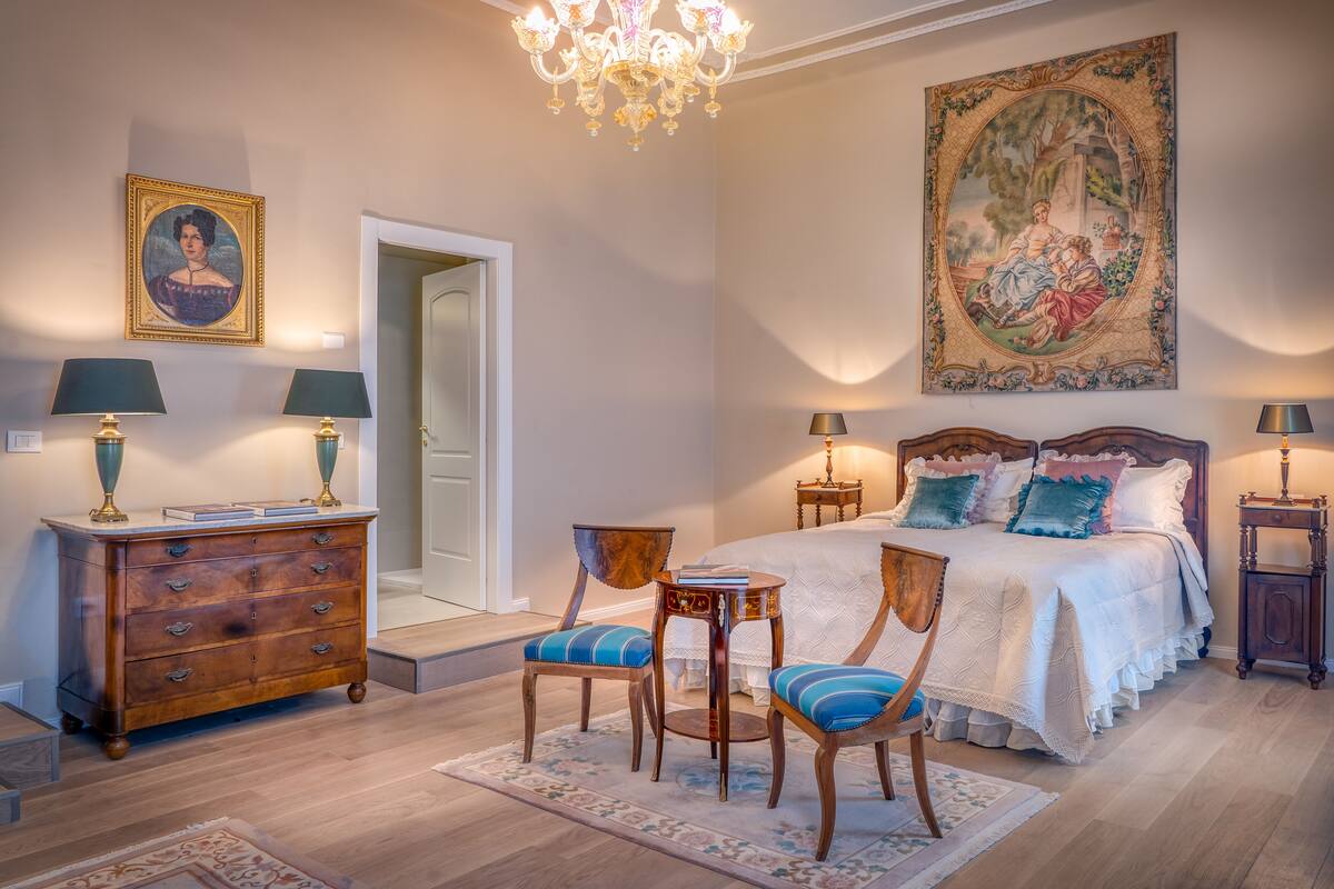 Quarto com cama, duas cadeiras, penteadeira, lustre e duas luminárias.