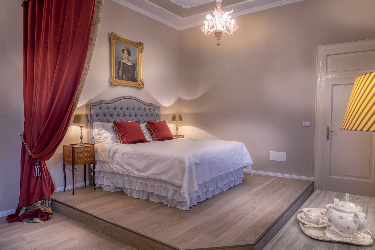 Quarto com cabeceira estofada, roupa de cama vermelha e branca, um candelabro e uma mesa de cabeceira com abajur.
