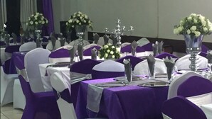 Salão de festas com mesas montadas para um evento formal, com toalhas roxas e cadeiras brancas com fitas roxas.