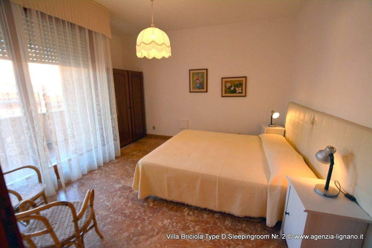 Quarto com cama, mesa de cabeceira com abajur, cadeira, guarda-roupa e duas imagens emolduradas na parede.