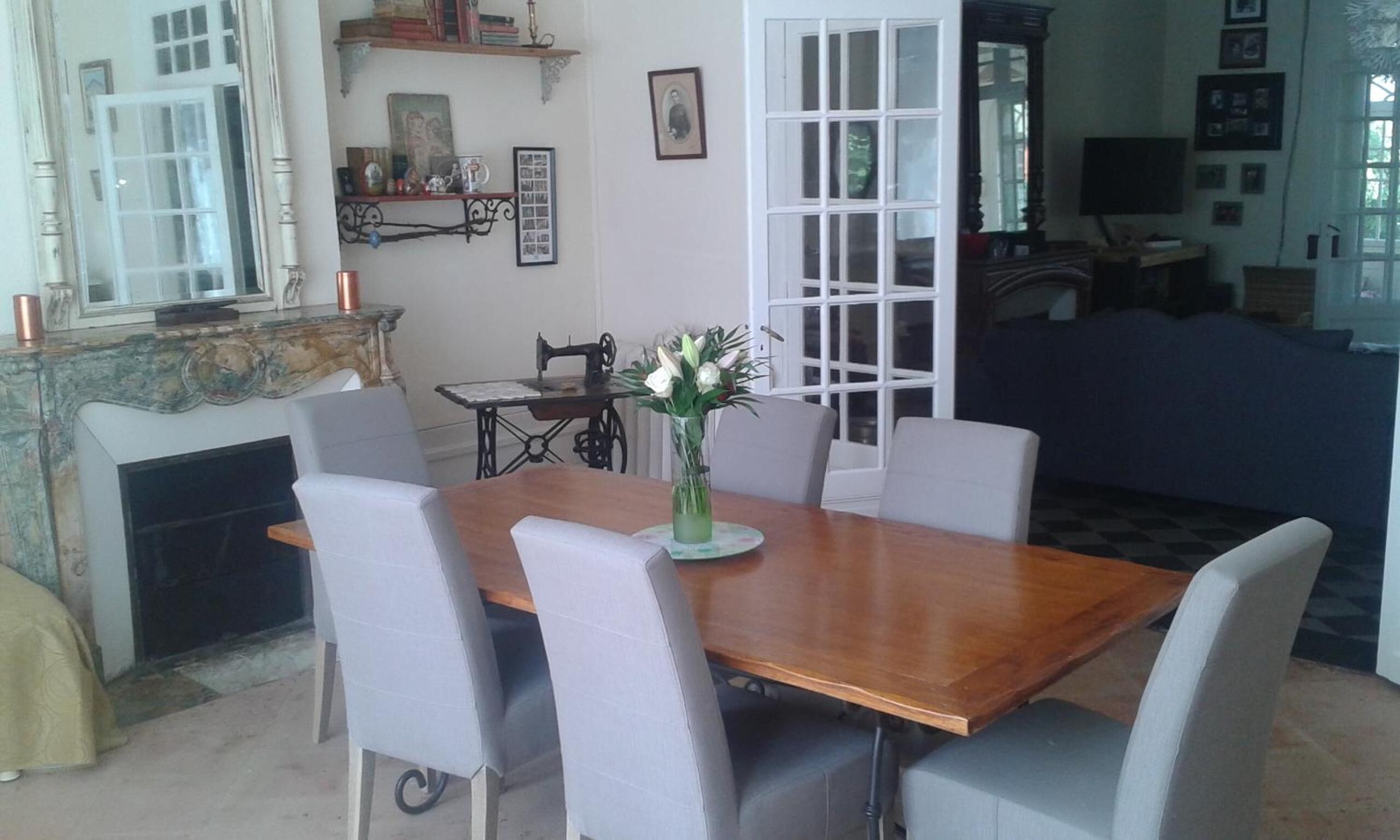 Uma sala de jantar com uma mesa de madeira, seis cadeiras, um vaso com flores e uma máquina de costura ao fundo.