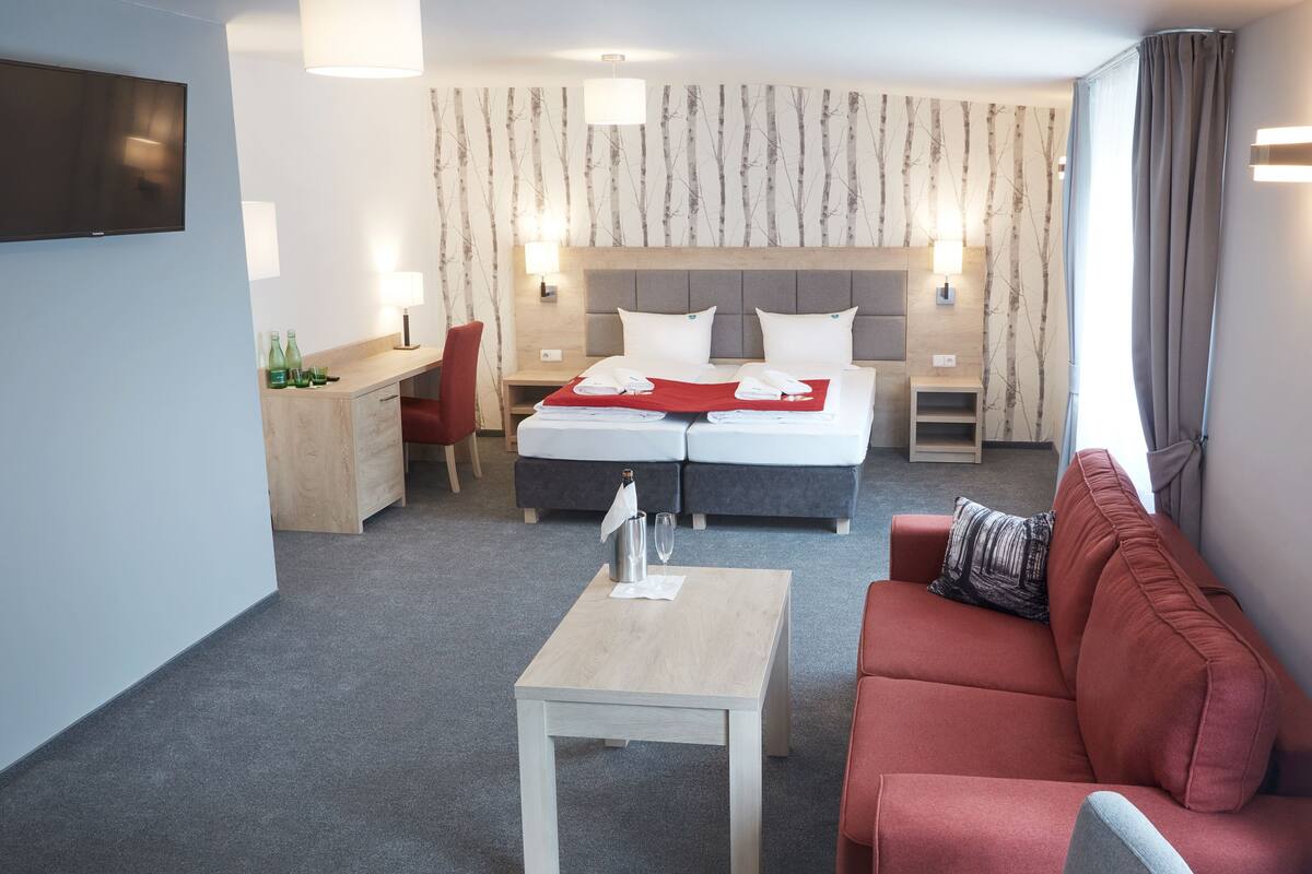 Quarto de hotel com cama, escrivaninha, sofá vermelho e televisão.