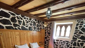 Quarto com parede de pedra, cabeceira de madeira, cama com manta colorida, mesa de cabeceira de madeira, cadeira de vime e janela com vista.