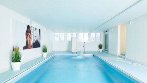 Uma grande piscina interior com água azul cristalina, rodeada por paredes brancas e plantas em vasos.