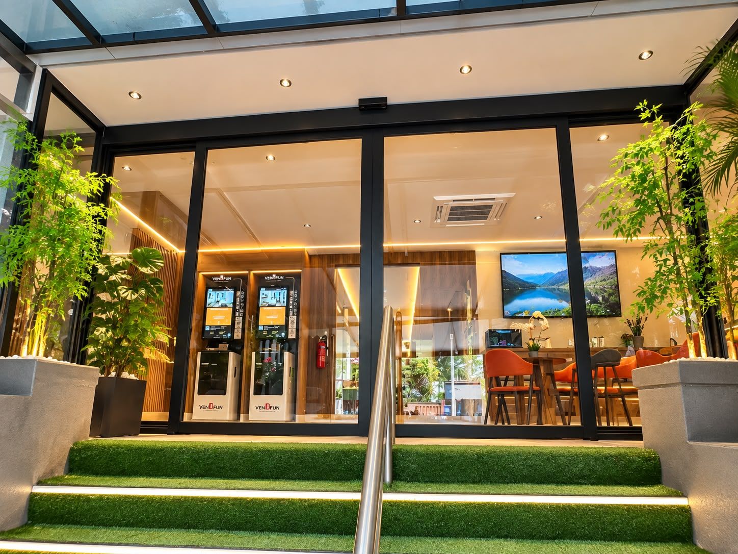 Um saguão de hotel moderno com entrada de vidro, plantas internas e escada que leva ao andar superior.