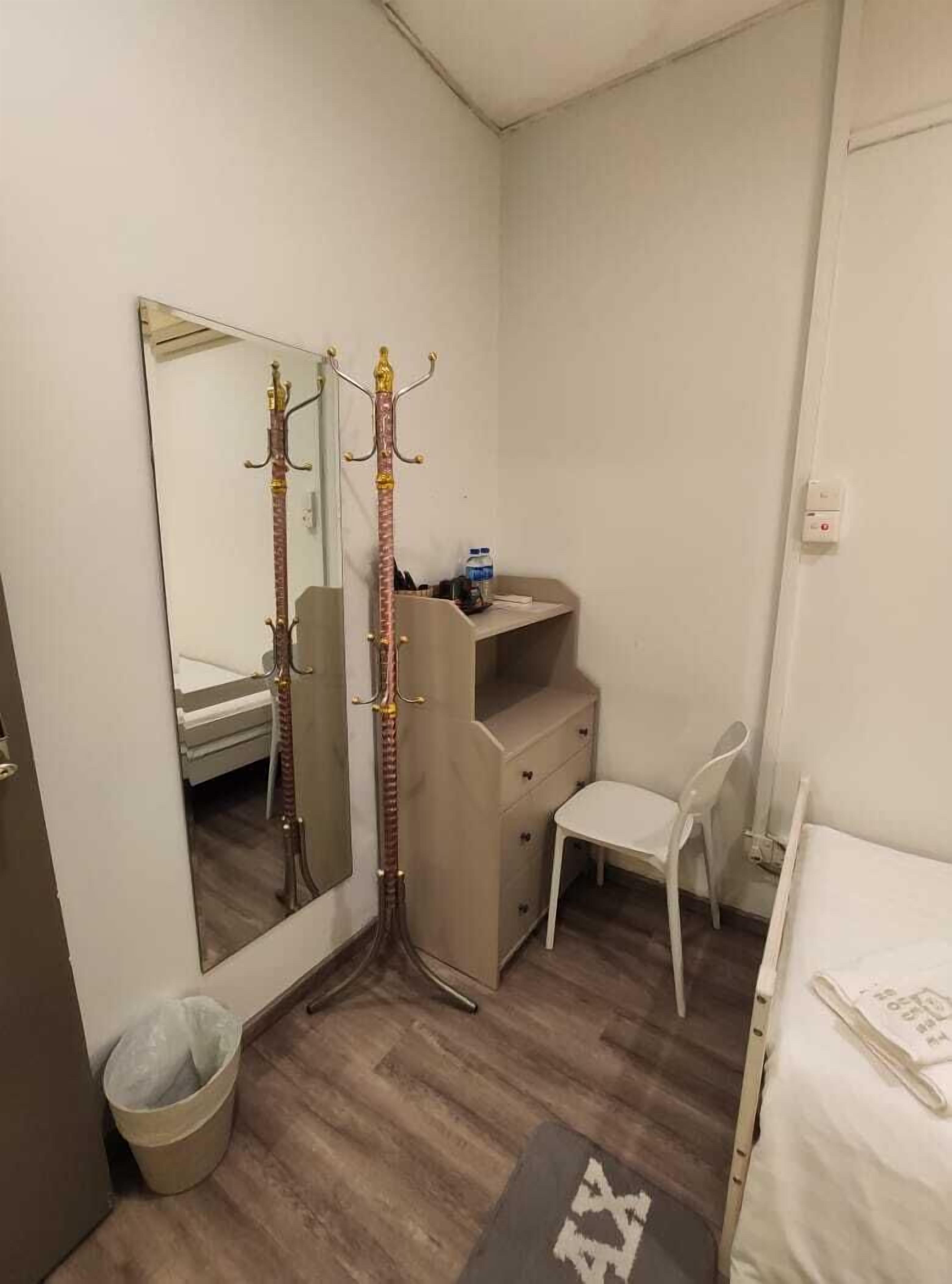 Quarto pequeno e bem iluminado, com piso de madeira, cama branca, escrivaninha com cadeira, espelho e cabideiro.