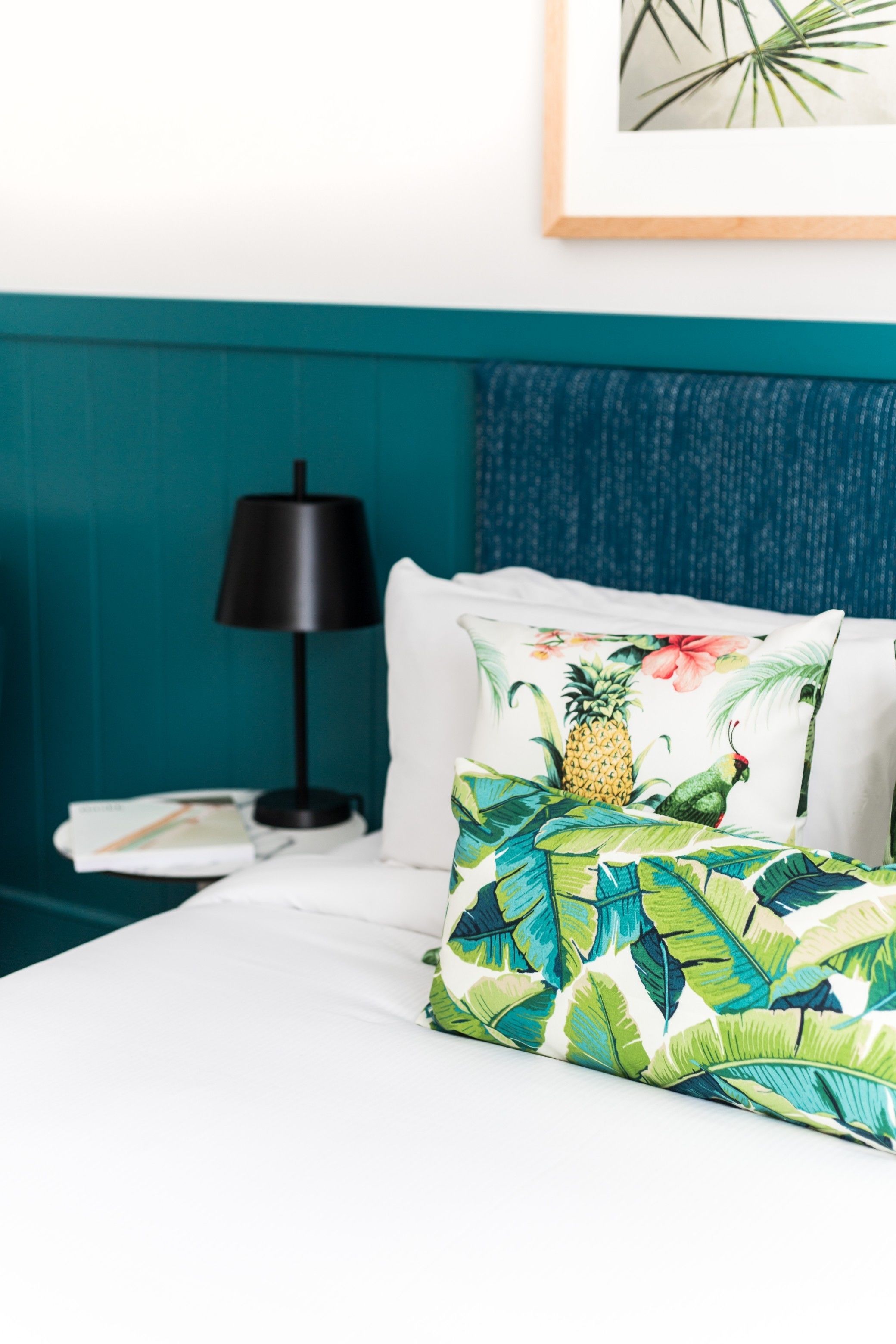 Cama bem arrumada com lençóis brancos, almofada com tema tropical, abajur preto e quadro na parede.