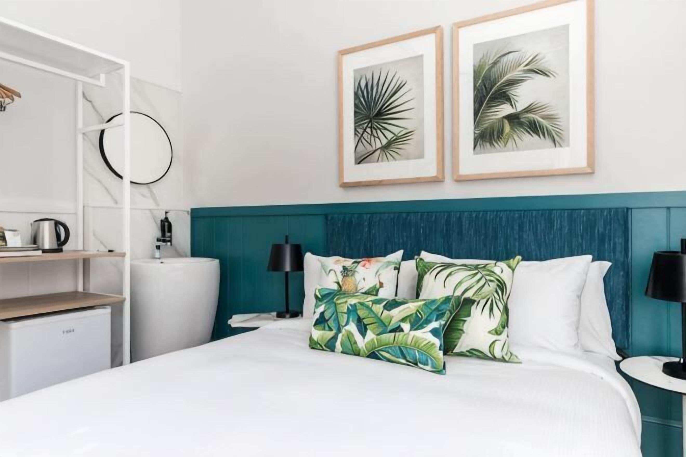 Quarto de hotel com cama, duas obras de arte com plantas emolduradas, cabeceira verde-petróleo e mesa de cabeceira com abajur.