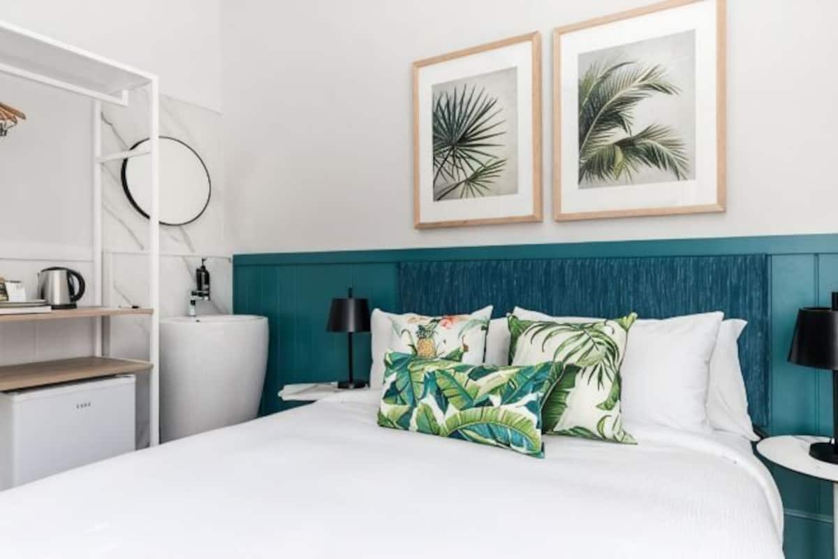 Quarto de hotel com cama, duas obras de arte com plantas emolduradas, cabeceira verde-petróleo e mesa de cabeceira com abajur.