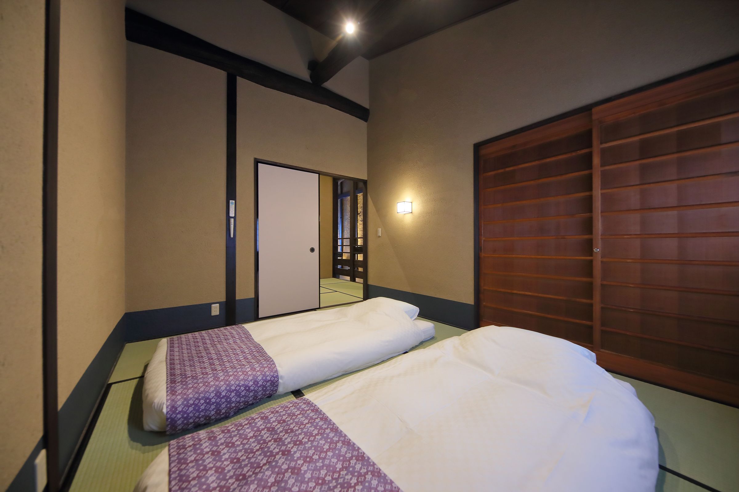 Quarto de hotel com duas camas, uma porta deslizante de madeira e uma pequena luminária fixada na parede.