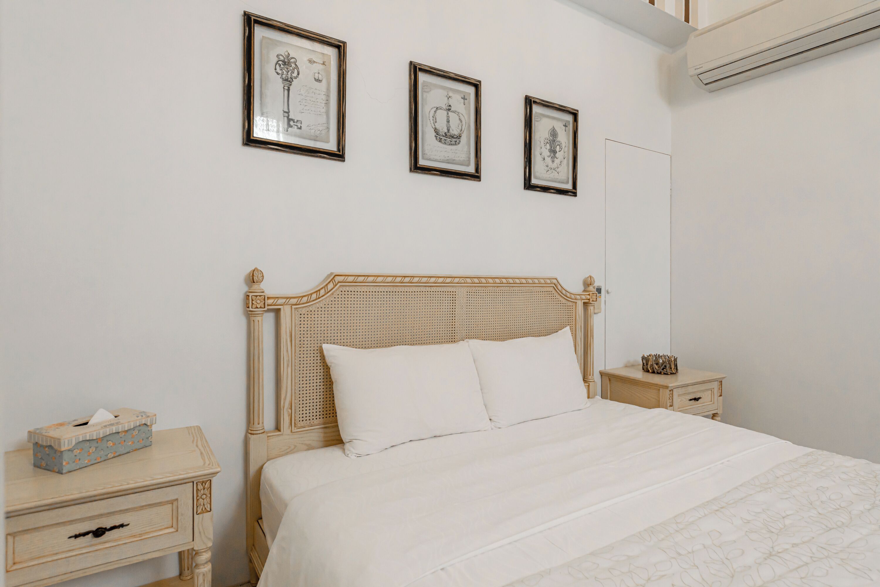 Uma cama bem arrumada com cabeceira de madeira, uma mesa de cabeceira com caixa de lenços e três quadros na parede.