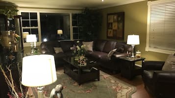 Uma sala de estar aconchegante com um sofá de couro escuro, uma mesa de centro de vidro e um tapete floral. Há duas luminárias em mesas laterais e uma janela grande com persianas.