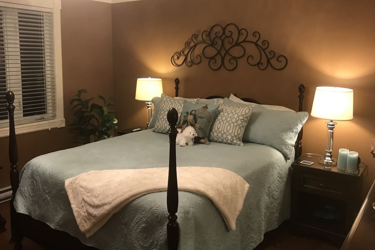 Quarto com cama, duas luminárias de cabeceira, um detalhe decorativo na parede e uma mesinha de cabeceira com abajur e velas.