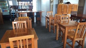 Interior de restaurante com mesas e cadeiras de madeira, área de bar ao fundo e chão coberto por carpete.