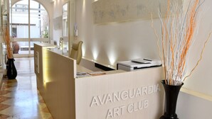 Balcão de recepção com uma placa indicando "AVANGUARDIA ART CLUB" em um interior moderno, com uma janela grande e uma planta decorativa.