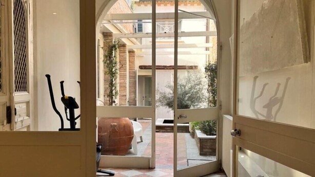 Um quarto amplo com piso xadrez, uma grande porta arqueada que dá para uma área externa com um barril e uma cadeira, e vista para um edifício com varanda.