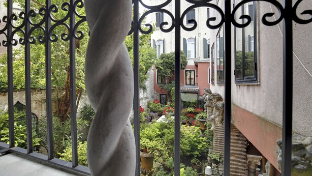 Visão através de grades de metal ornamentadas, revelando um pátio com plantas em vasos, uma estátua e um edifício com várias janelas.