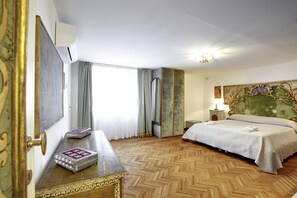 Quarto de hotel com piso de madeira em padrão herringbone, cama com colcha branca, mesa de cabeceira com uma caixa decorativa, um espelho grande e ar condicionado fixado na parede.