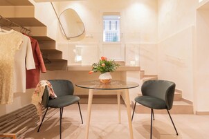 Quarto moderno com mesa de vidro, duas cadeiras, um vaso com flores, um cabideiro com roupas e uma janela com vista para o exterior.