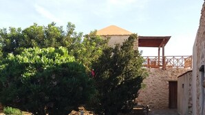 Uma trilha de pedra leva a um edifício com área coberta, rodeado por vegetação e muros de pedra.