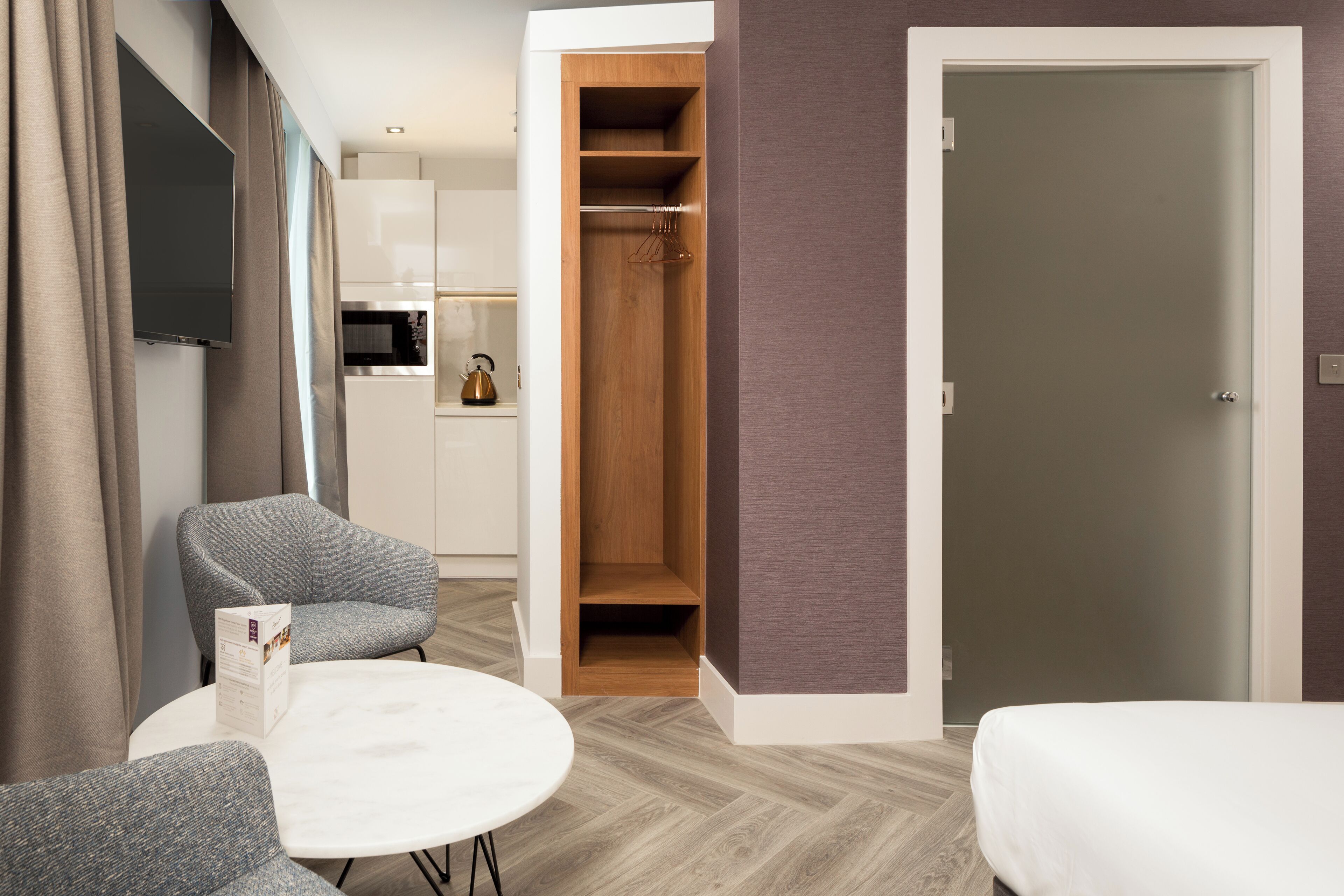 Quarto de hotel moderno com uma poltrona cinza, uma pequena mesa redonda, um guarda-roupa e uma porta.