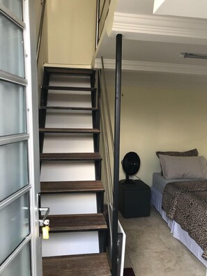 Escada de madeira com corrimãos de metal que leva a um quarto com cama e mesa de cabeceira pequena.