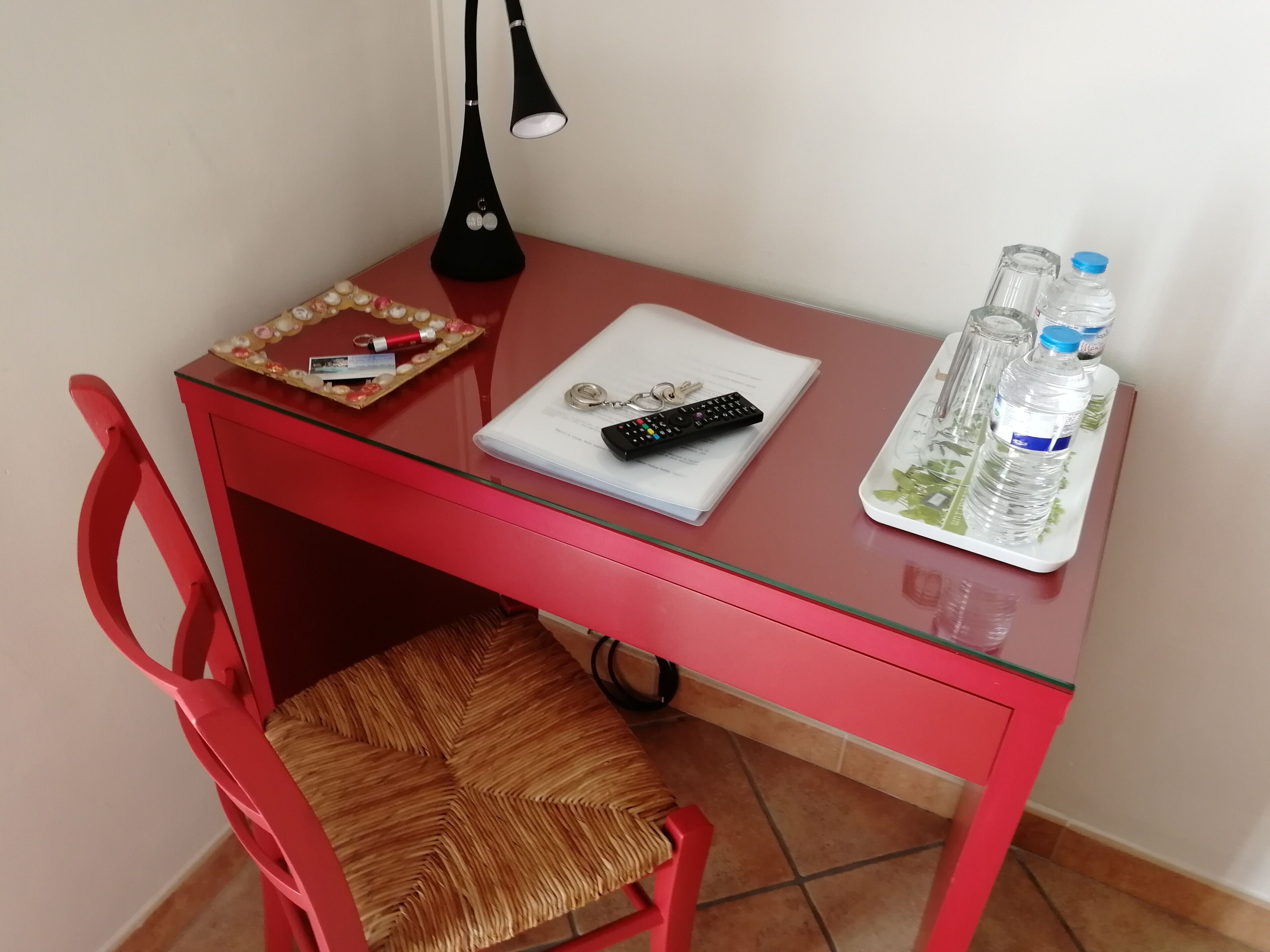 Uma mesa vermelha com tampo de vidro, uma luminária de mesa preta, um controle remoto, uma bandeja com chaves e uma garrafa de água.
