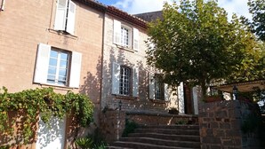 Um edifício de pedra com escadaria que leva à entrada, rodeado por vegetação e uma árvore.
