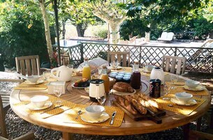 Mesa de madeira redonda posta para o pequeno-almoço, com pratos, chávenas e diversas iguarias.