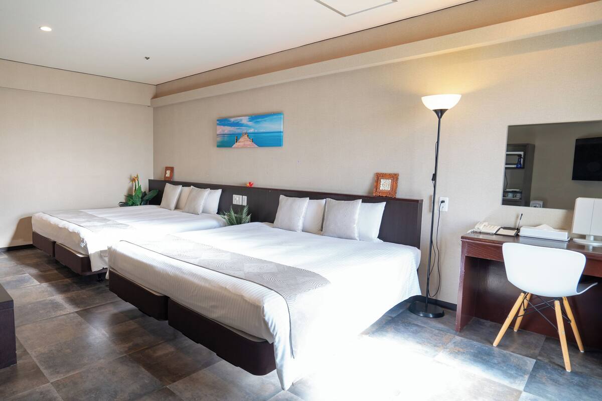 Quarto de hotel com duas camas, uma escrivaninha, uma cadeira, uma luminária de mesa e um quadro na parede.