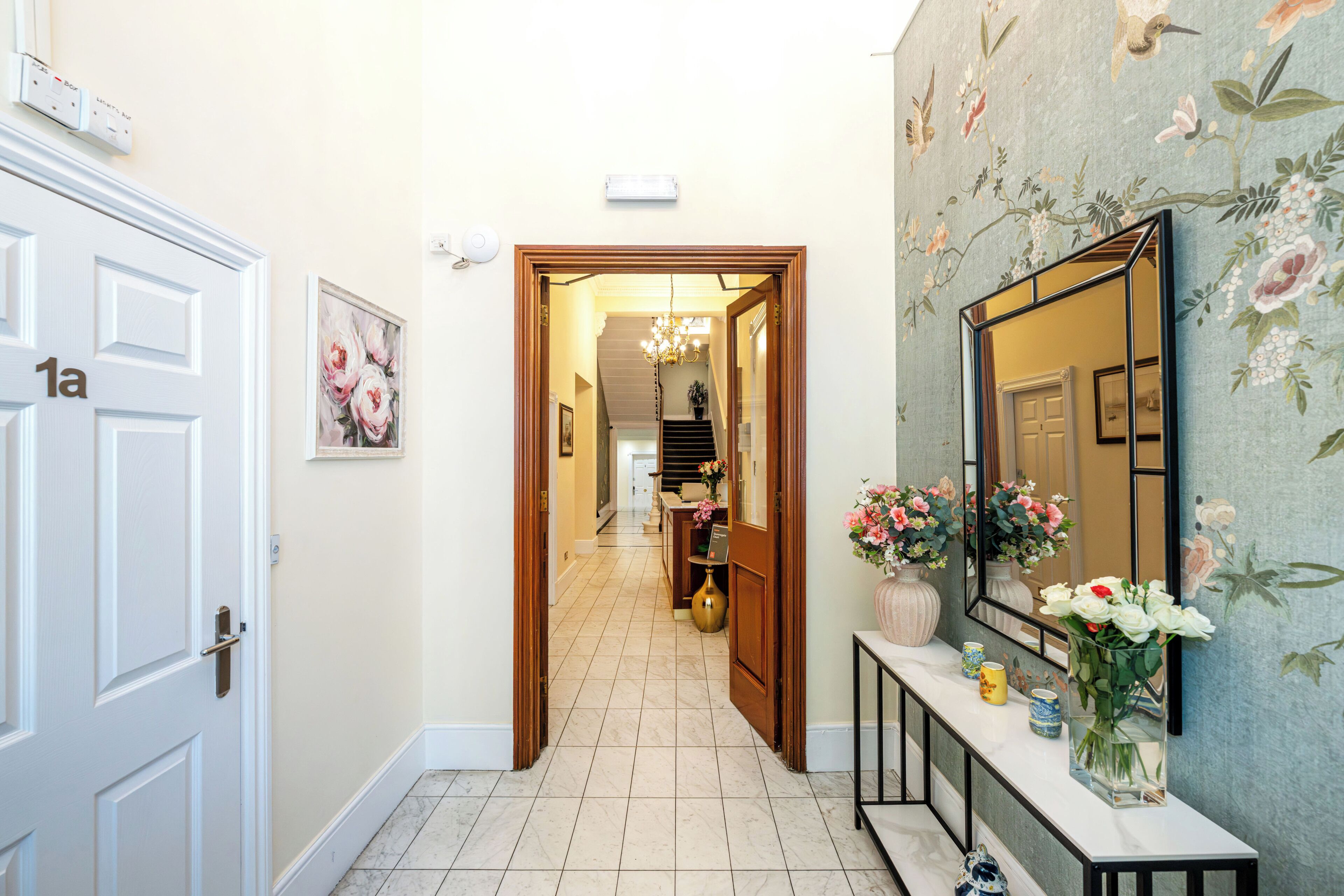 Um corredor com uma porta identificada como '1a', um quadro emoldurado, uma mesa consola com um vaso de flores e um espelho na parede.