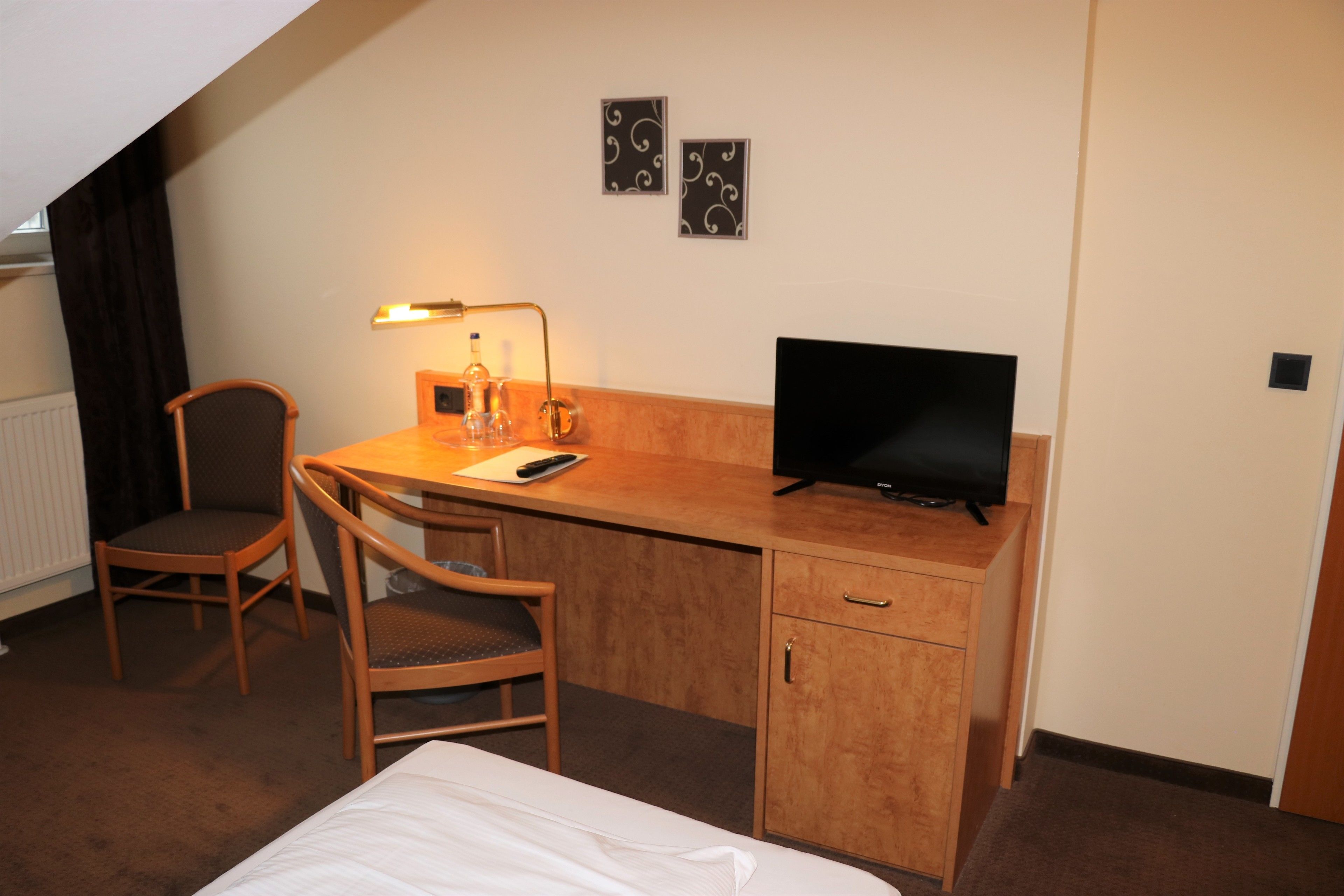 Quarto de hotel com uma cama, uma escrivaninha com computador, uma cadeira, um abajur e duas obras de arte nas paredes.