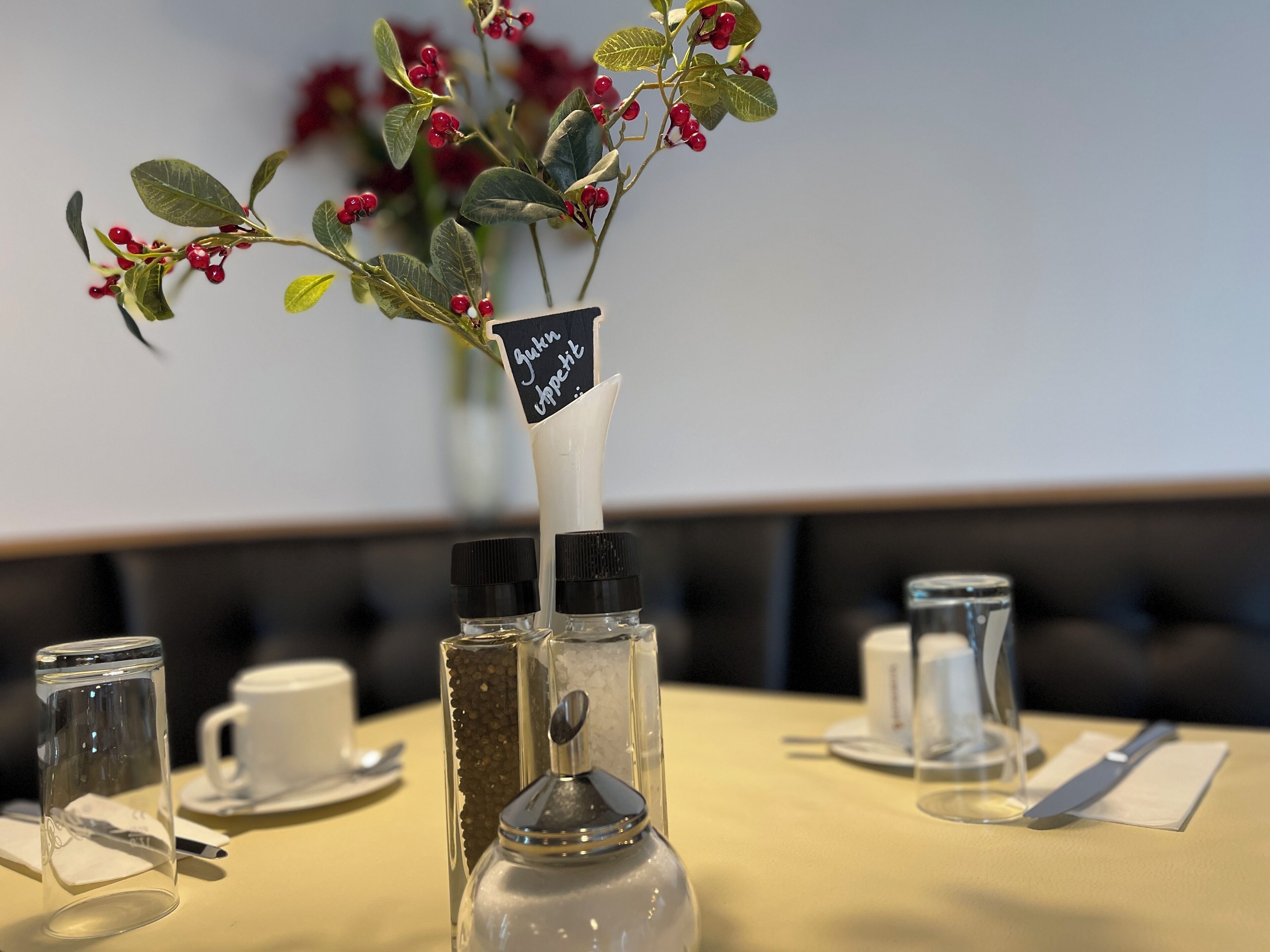 Uma mesa posta com um jarro de vidro transparente, uma placa preta pequena e um galho decorativo com berris vermelhas.