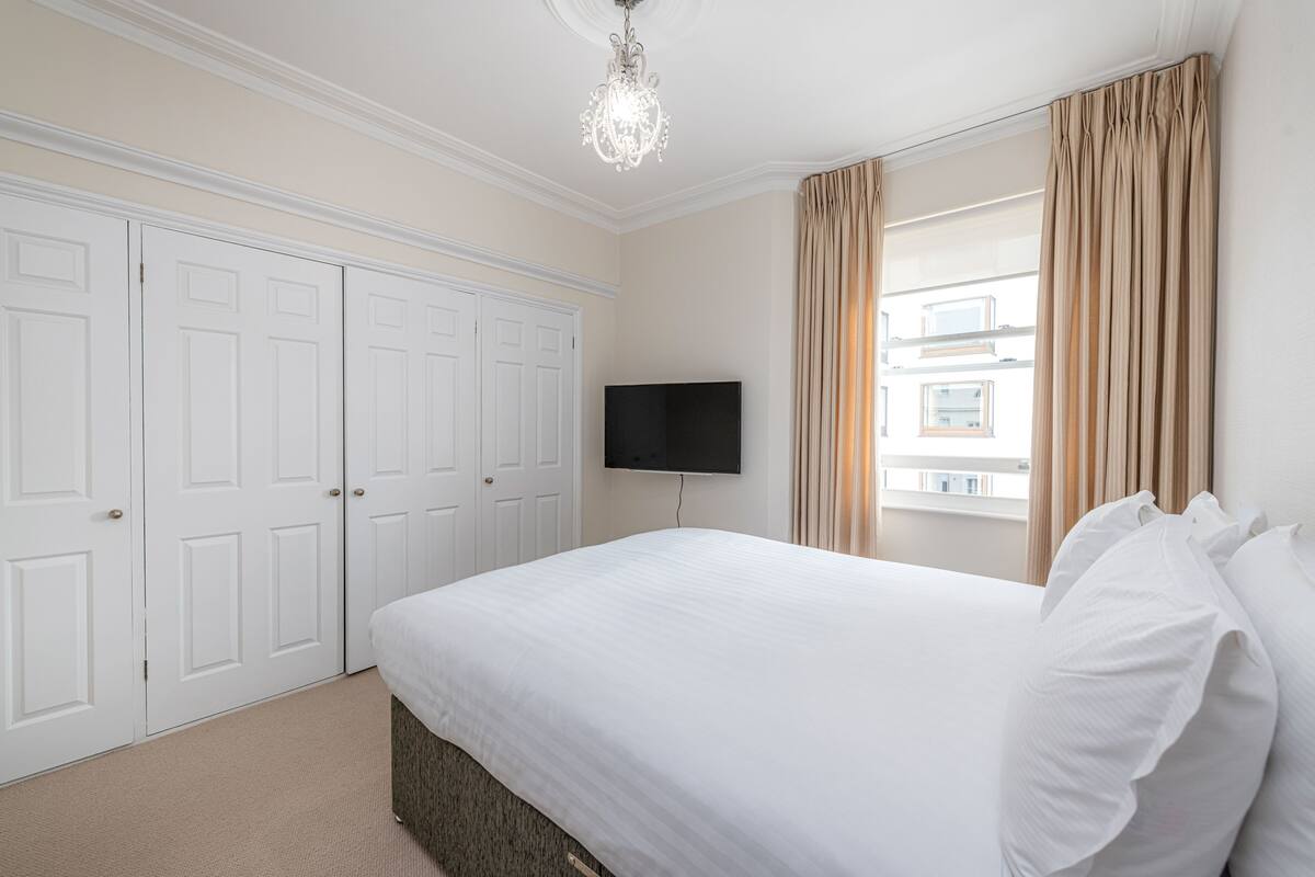 Quarto com cama grande, televisão de tela plana, lustre e vista para o exterior.