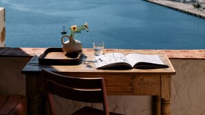 Uma mesa com um vaso, um livro e um copo, com vista para a costa e um arco de pedra.