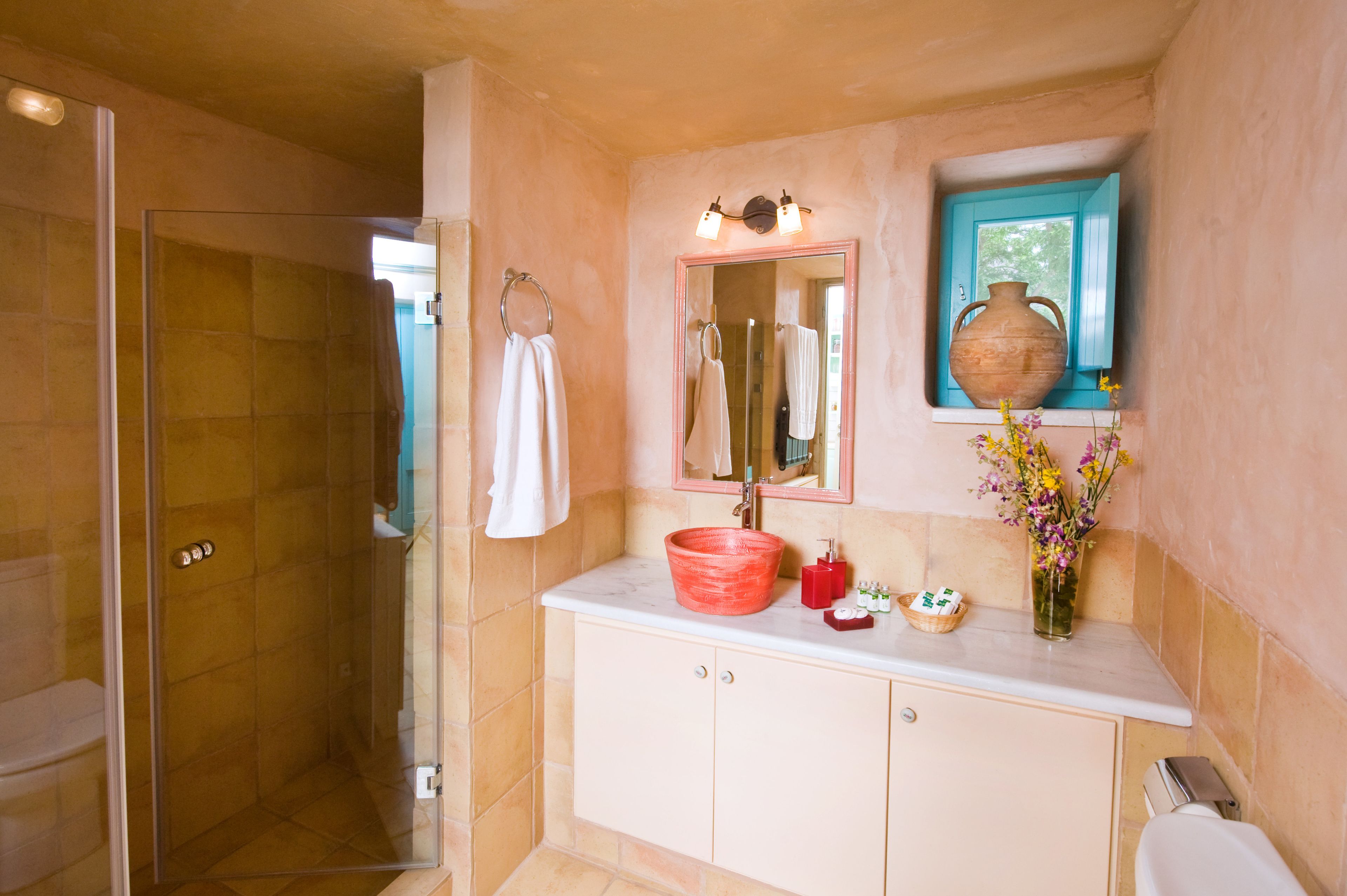 Casa de banho com cabine de duche de vidro, lavatório branco com cuba vermelha e um vaso de flores na toucador.