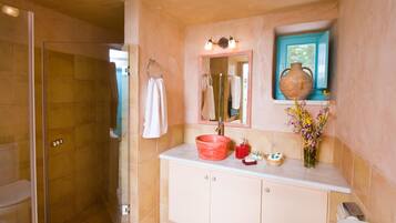 Casa de banho com cabine de duche de vidro, lavatório branco com cuba vermelha e um vaso de flores na toucador.
