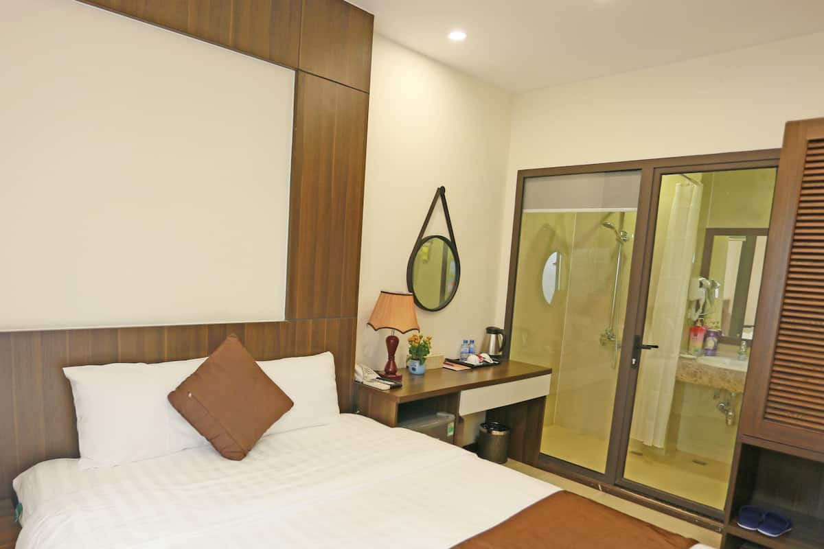 Quarto de hotel com cama, mesa de cabeceira, luminária e porta de vidro deslizante para uma área de chuveiro.