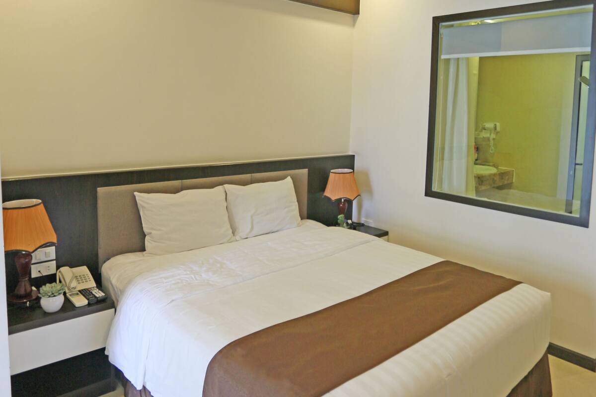 Quarto de hotel com uma cama grande, mesa de cabeceira com abajur, telefone e uma pequena mesa com planta.