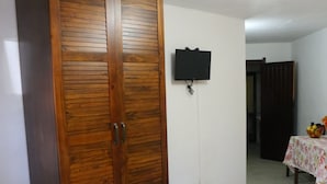 Um quarto com um guarda-roupa de madeira grande, uma cama com colcha estampada, uma televisão fixada na parede e uma mesinha de cabeceira com uma fruteira.