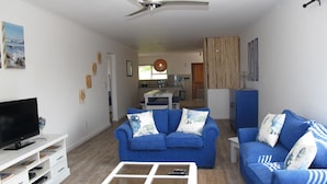 Uma sala de estar com televisão, dois sofás azuis, uma mesa de centro branca e um ventilador de teto.