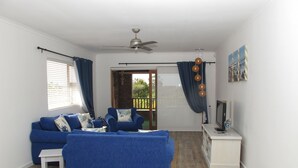 Sala de estar com sofá azul, mesinha lateral branca, ventilador de teto e vista para o exterior através de uma porta de vidro.