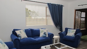 Uma sala de estar com um sofá azul, uma mesa de centro de vidro, uma janela com persianas e cortinas.
