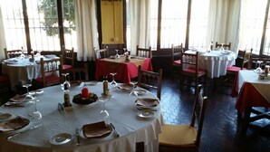 Uma área de refeições com mesas redondas para quatro pessoas, cada uma com toalhas brancas, guardanapos vermelhos e copos de vinho.