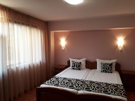 Cama arrumada com colcha estampada, duas luminárias de cabeceira e uma janela com cortinas finas.