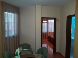 Quarto de hotel com mesa de jantar, duas cadeiras, sofá verde, cama com cobertura estampada e banheiro visível por uma porta.